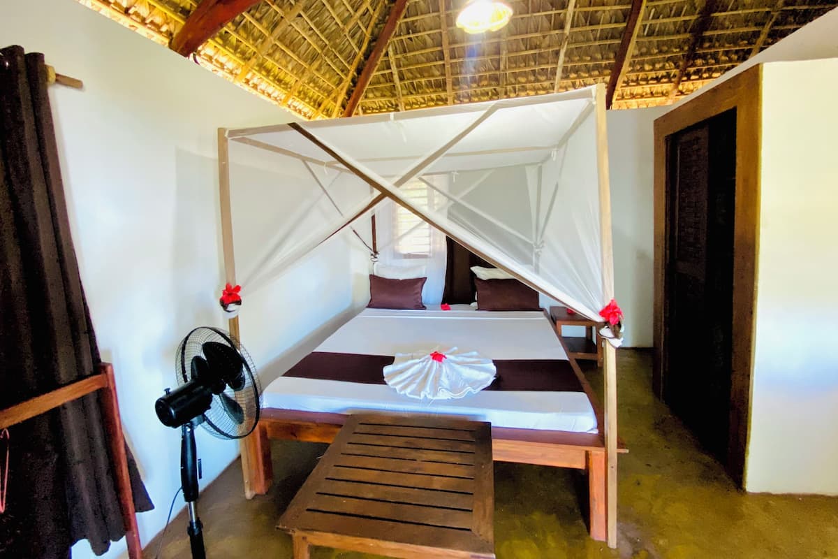 Quarto com cama sob uma rede mosquiteira, um ventilador e uma mesa de madeira.