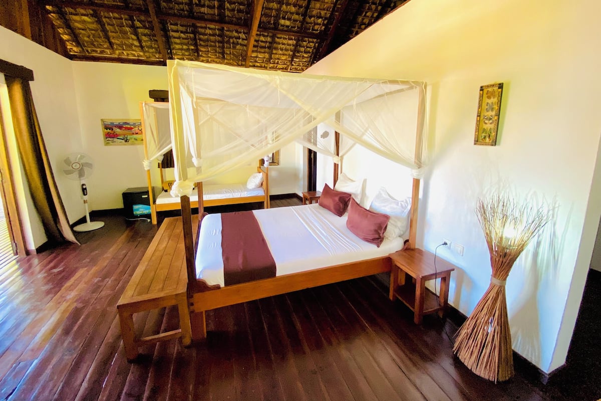Quarto com cama de madeira, mosquiteiro, mesa de cabeceira, ventoinha e um vaso decorativo.