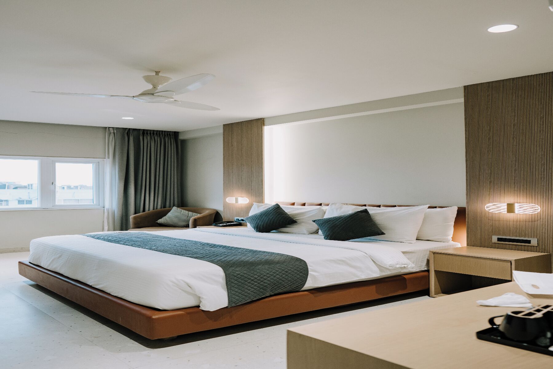 Quarto de hotel moderno com uma cama grande, escrivaninha, cadeira, ventoinha no teto e janela com cortinas.