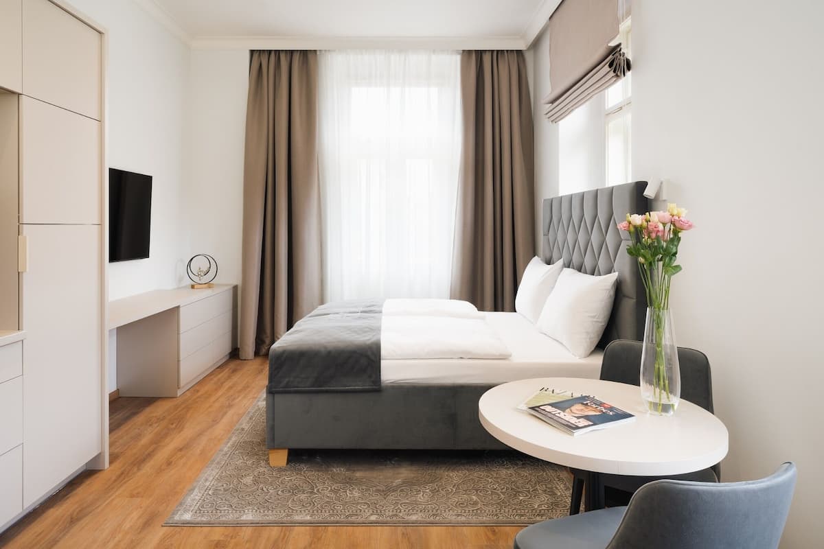 Quarto de hotel com cama, mesa redonda, televisão, escrivaninha e janela com cortinas.