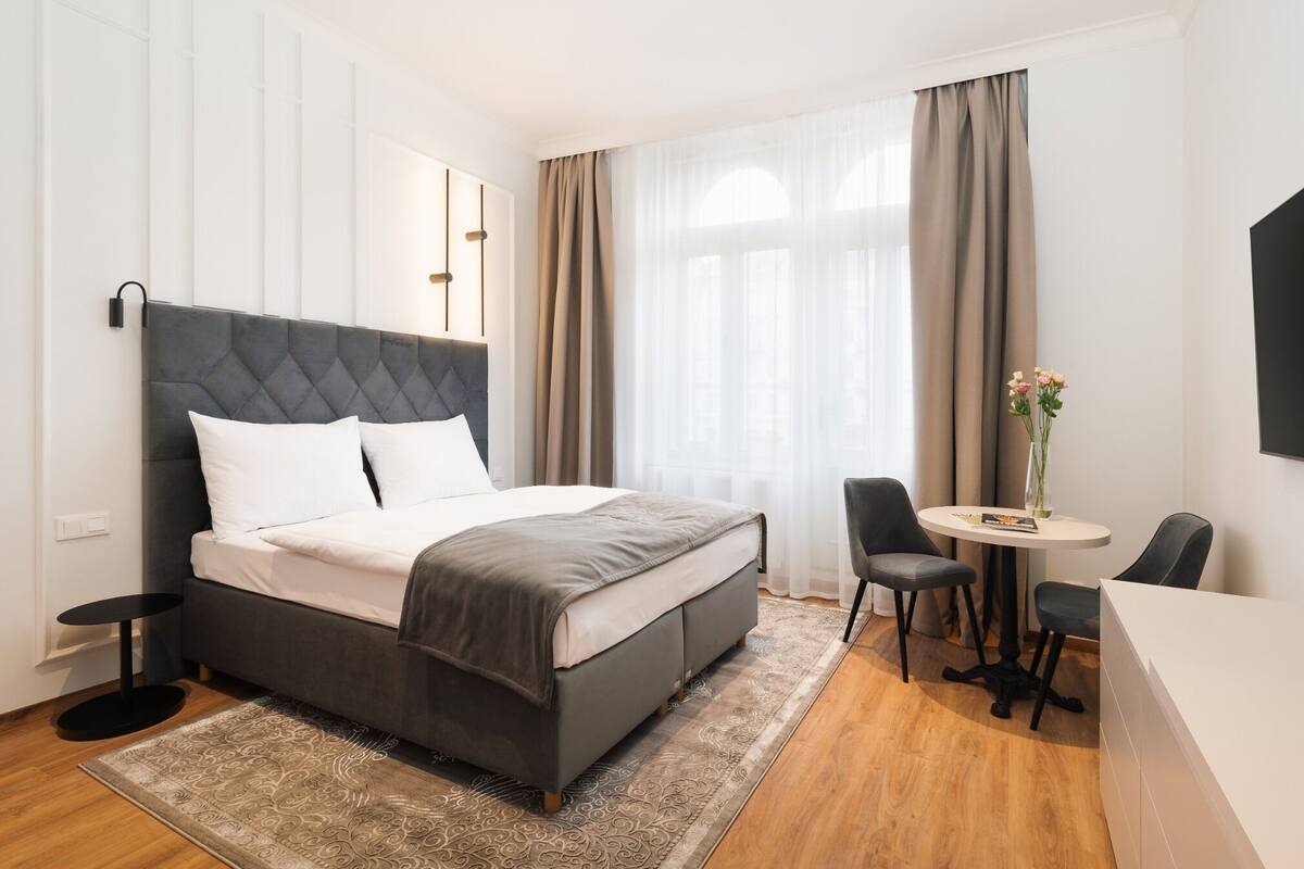Quarto de hotel moderno com uma cama grande, uma mesinha redonda, duas cadeiras e uma escrivaninha.