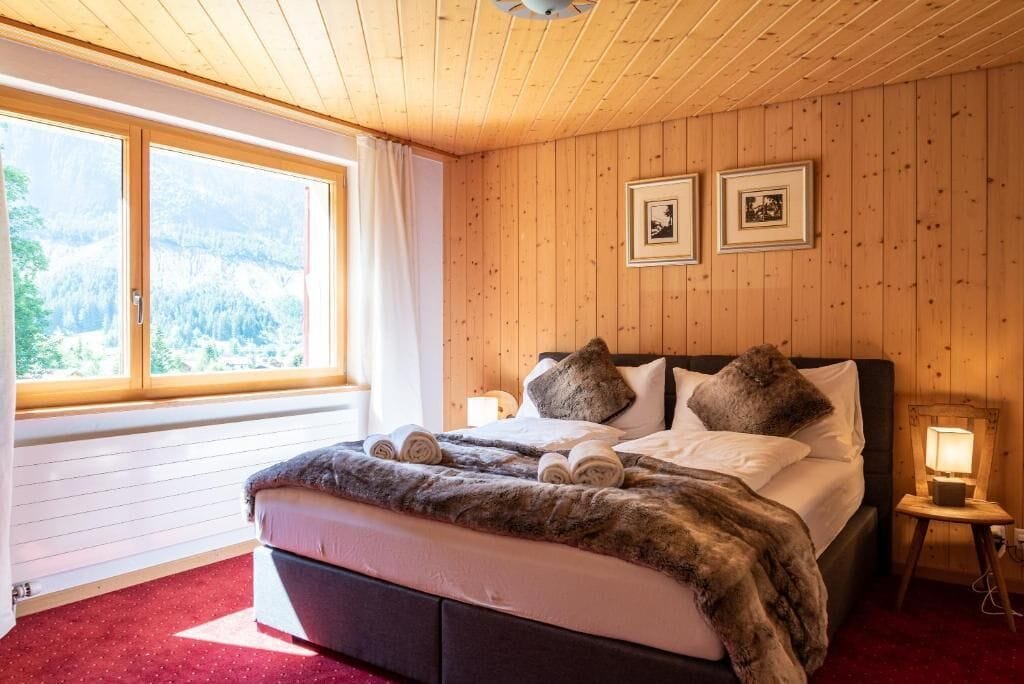 Um quarto com parede revestida de madeira, uma janela grande com cortinas, uma cama com manta de pele e duas imagens emolduradas na parede.