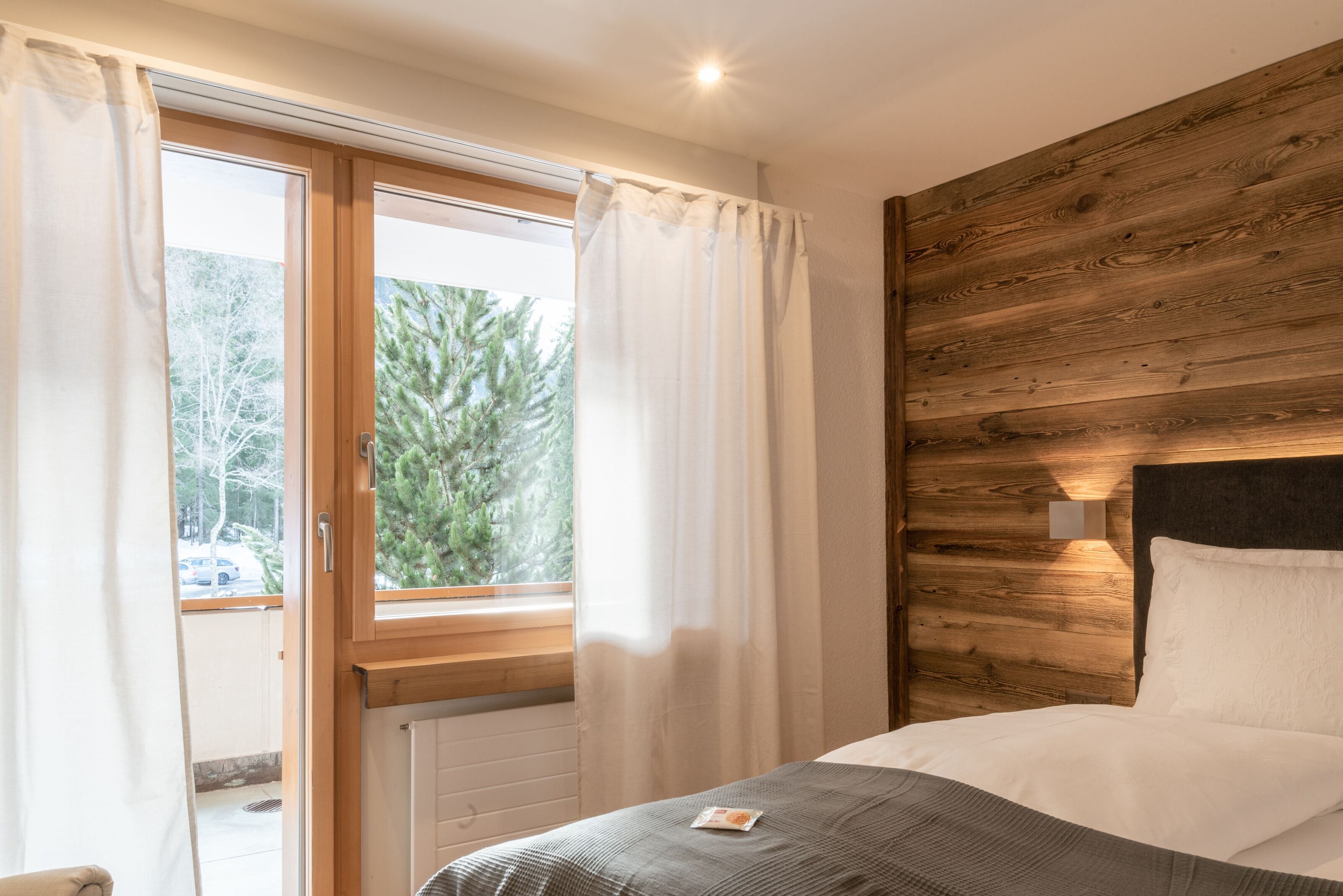 Quarto com cabeceira de madeira, cama com roupa de cama branca, janela com cortinas e vista para árvores lá fora.