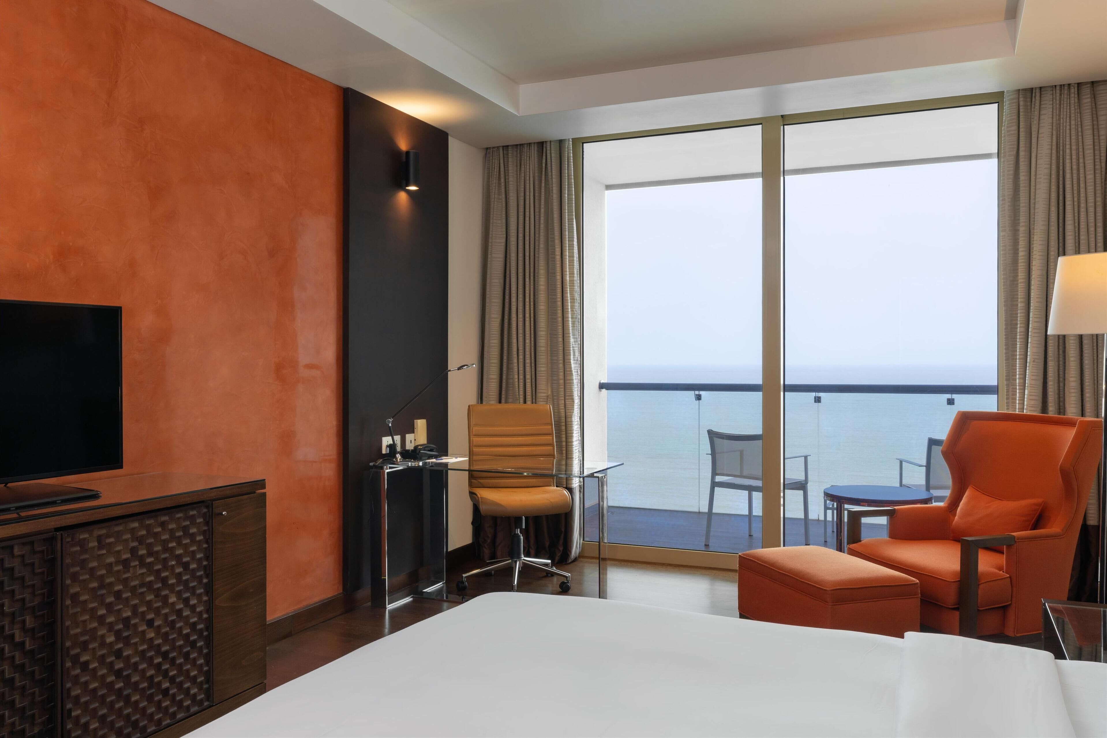 Quarto de hotel com uma cama grande, televisão, escrivaninha com cadeira e varanda com vista para o mar.