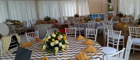 Salão de festas com mesas redondas cobertas por toalhas brancas, cadeiras brancas e guardanapos dourados.