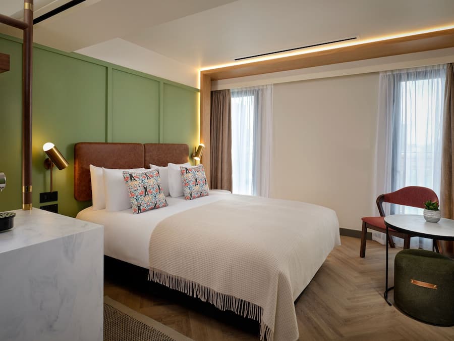 Quarto de hotel com uma cama grande, uma escrivaninha com luminária, uma cadeira e uma pequena mesa com uma planta.