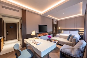Quarto de hotel moderno com televisão de tela plana, lareira, cama, escrivaninha com luminária e área de estar.