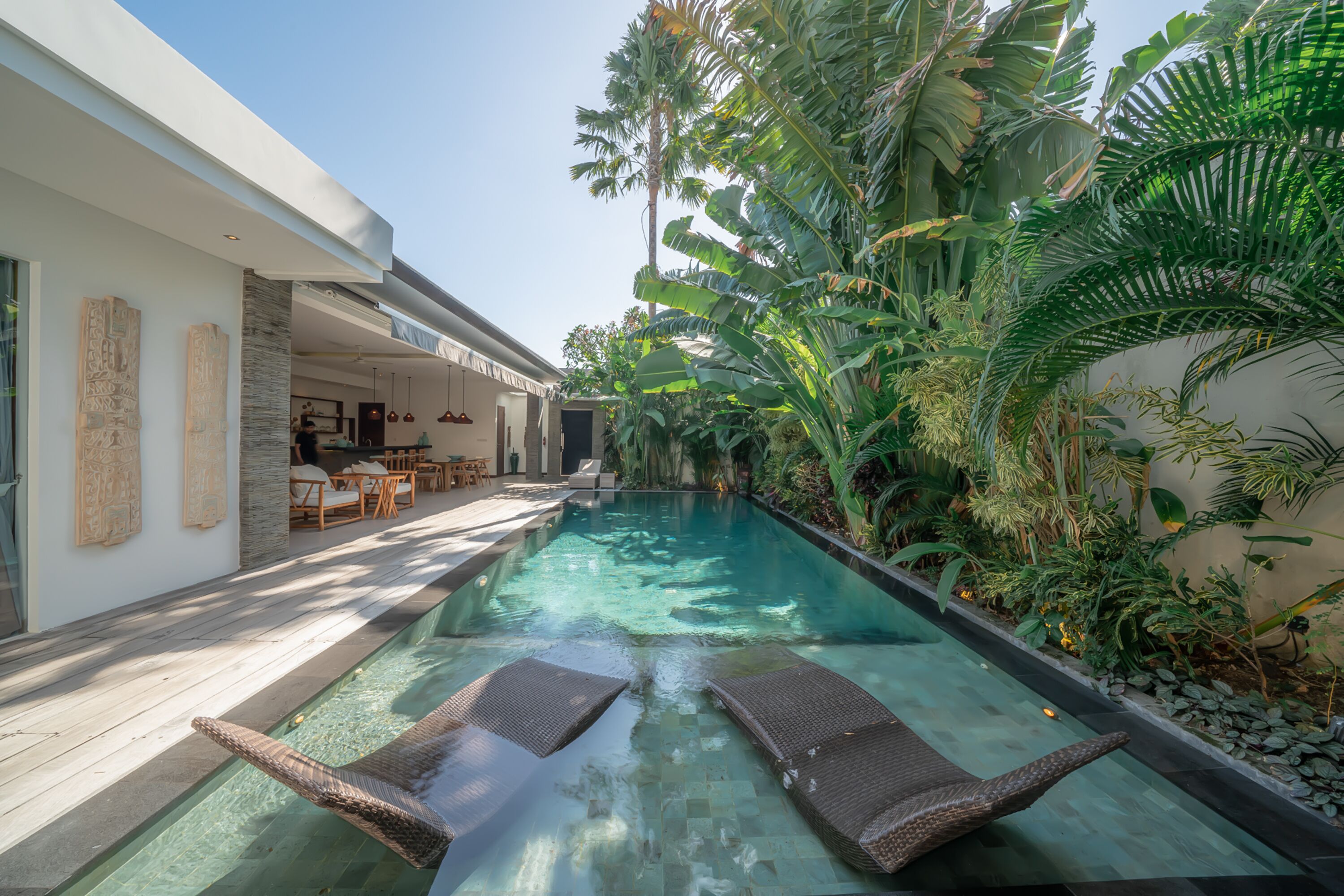 Uma área de piscina com espreguiçadeiras, espaço para refeições e vegetação tropical exuberante.