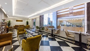 Um lobby moderno de hotel com piso xadrez, assentos confortáveis, área de refeições e vista da cidade através de grandes janelas.