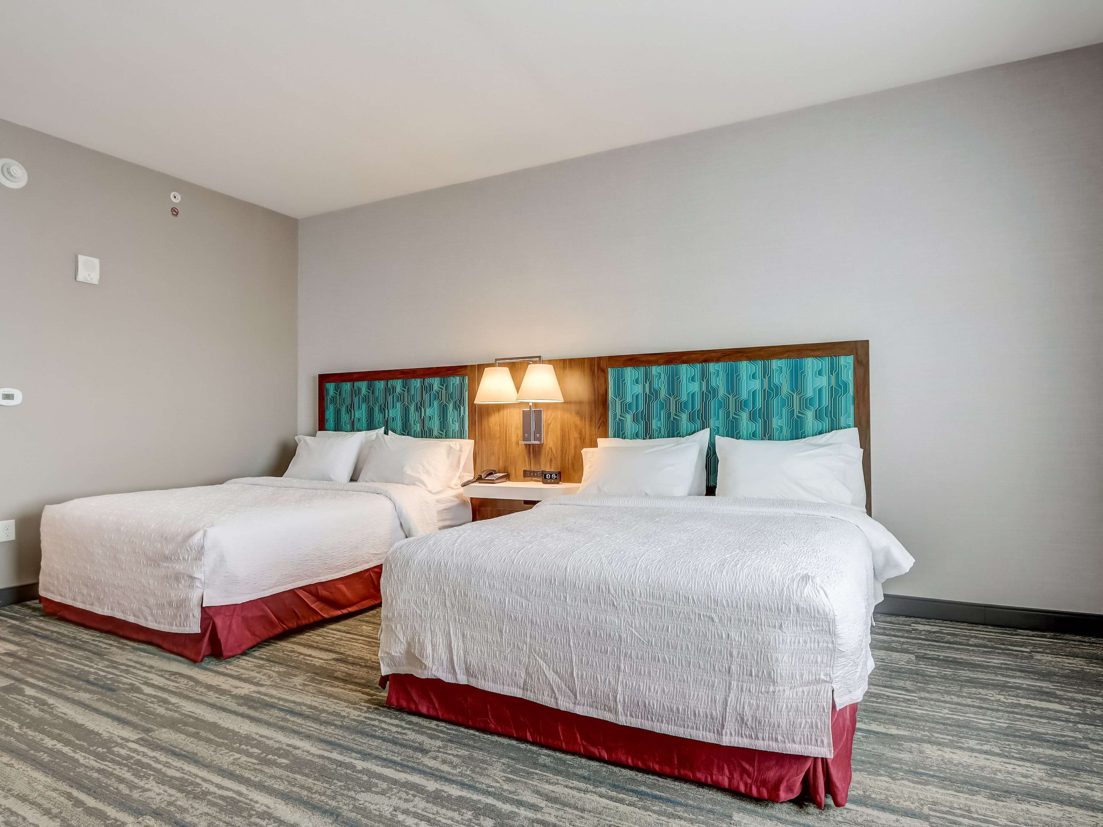 Quarto de hotel com duas camas, uma mesa de cabeceira, uma luminária e um termostato fixado na parede.