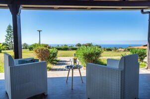 Área de estar coberta ao ar livre com móveis de vime e uma mesa posta para dois, com vista para um jardim paisagístico e o oceano.