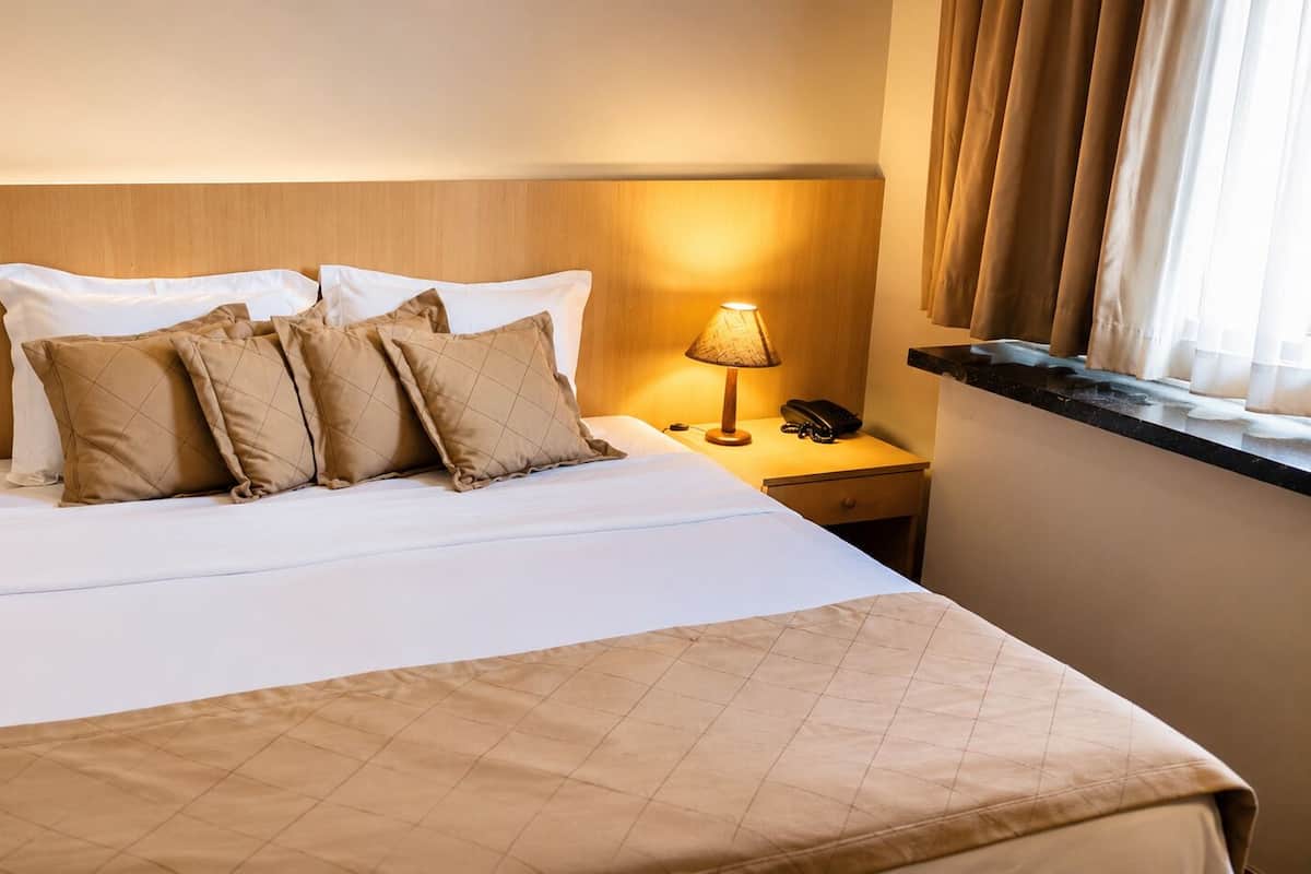 Quarto de hotel com cama, abajur, telefone e janela com cortinas.