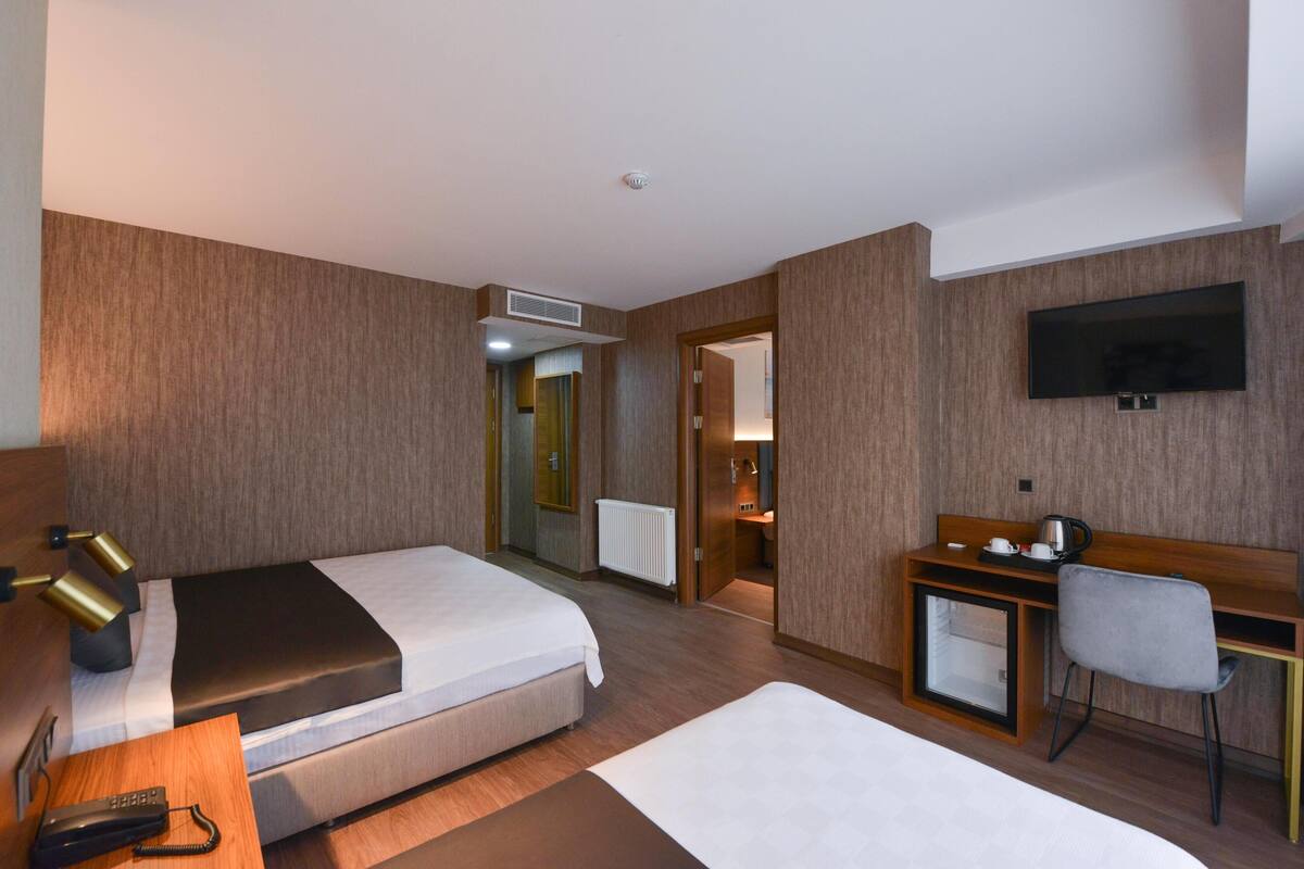 Quarto de hotel com duas camas, uma escrivaninha com cadeira, televisão na parede e uma pequena área de kitchenette.