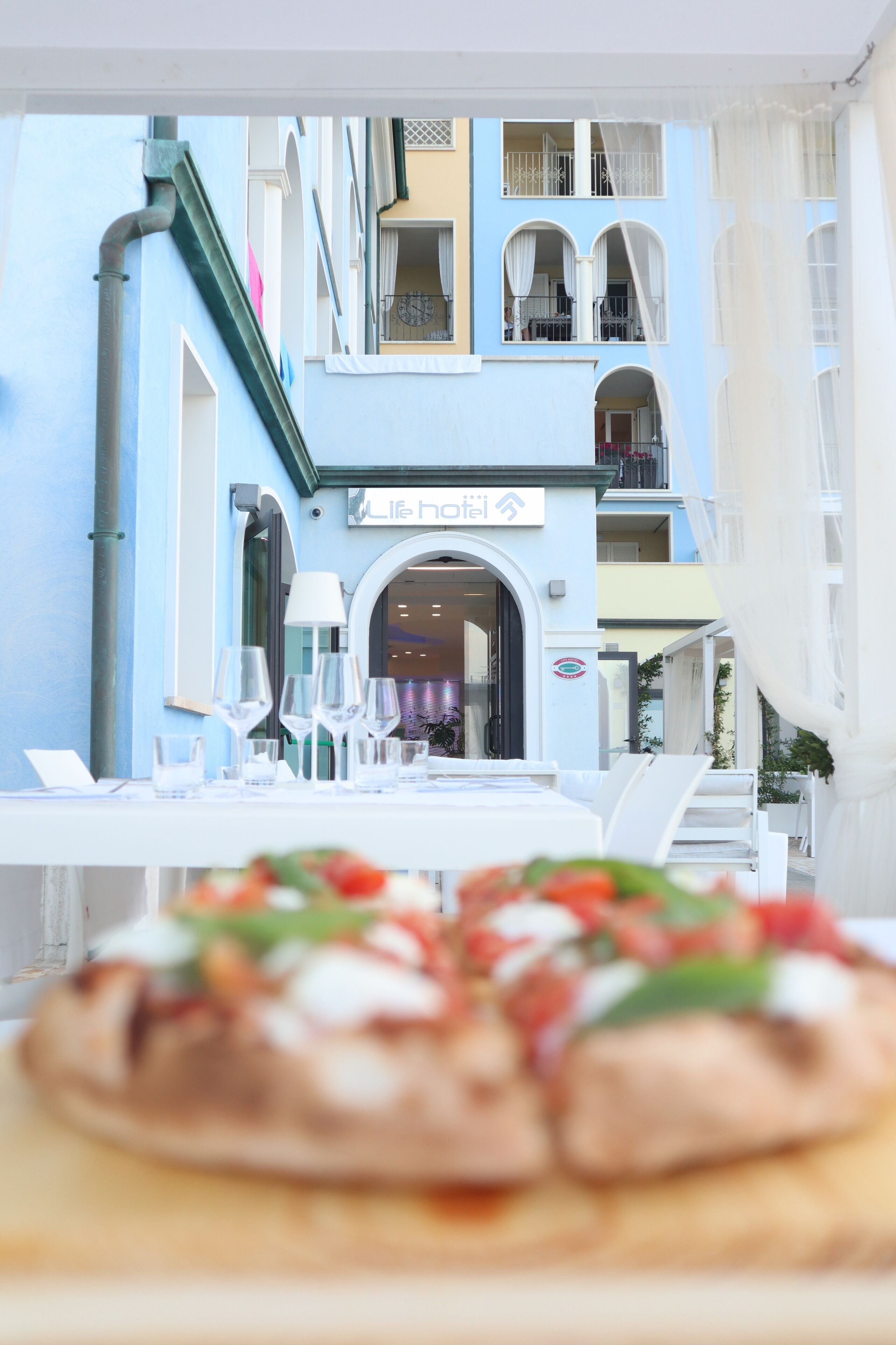 Restaurante com mesas ao ar livre, um edifício azul ao fundo e uma pizza em primeiro plano.