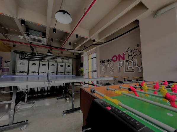 Um quarto com uma mesa de futebol de mesa, uma mesa comprida com toalha branca e uma parede com a inscrição 'Game ON LET'S PLAY'.