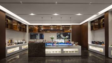 Um hall de hotel moderno com balcão de recepção de mármore, estantes de madeira e diversas comodidades, incluindo um espaço para café e plantas decorativas.