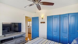 Quarto com cama, televisão sobre um móvel, ventoinha no teto e portas azuis.