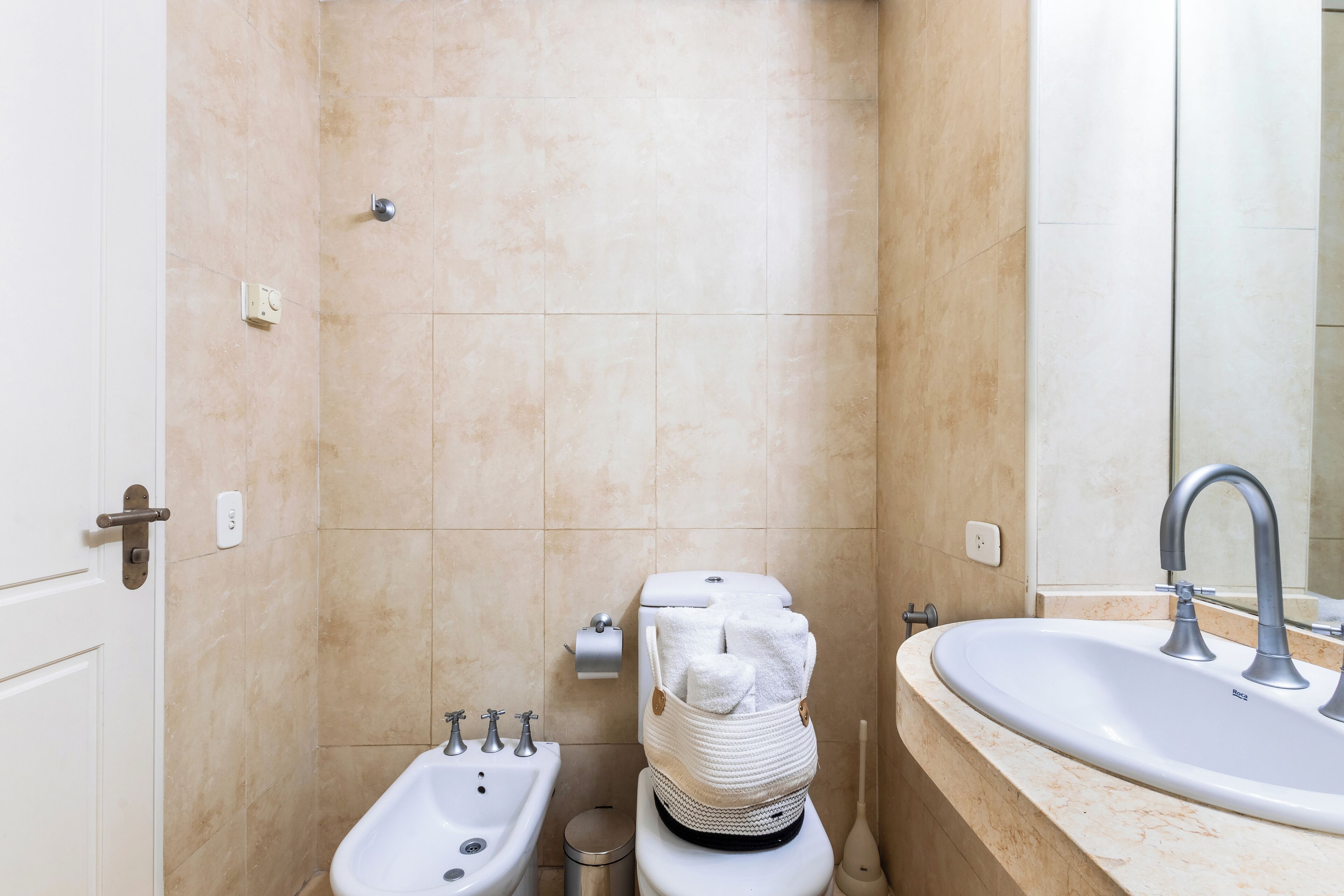 Casa de banho com azulejos bege, uma pia branca, um bidé e uma sanita com suporte para toalha.
