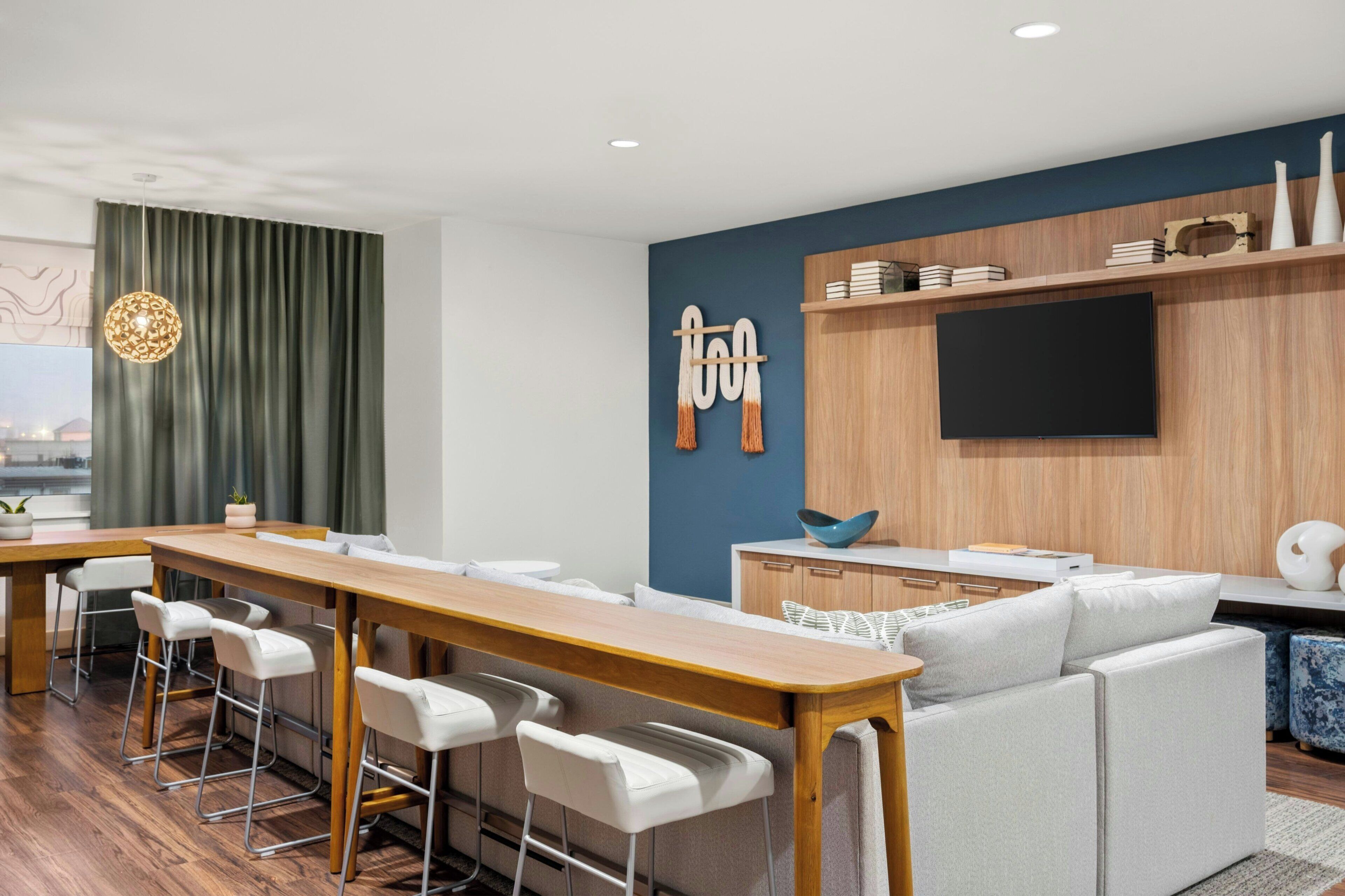Uma sala de estar moderna com uma mesa de jantar de madeira, banquetas brancas, um sofá cinza e uma TV de tela plana fixada em uma parede de madeira.