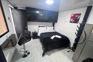 Quarto de hotel compacto com uma cama, uma secretária pequena com cadeira, uma TV fixada acima da cama, um cabideiro e uma parede com um quadro emoldurado.