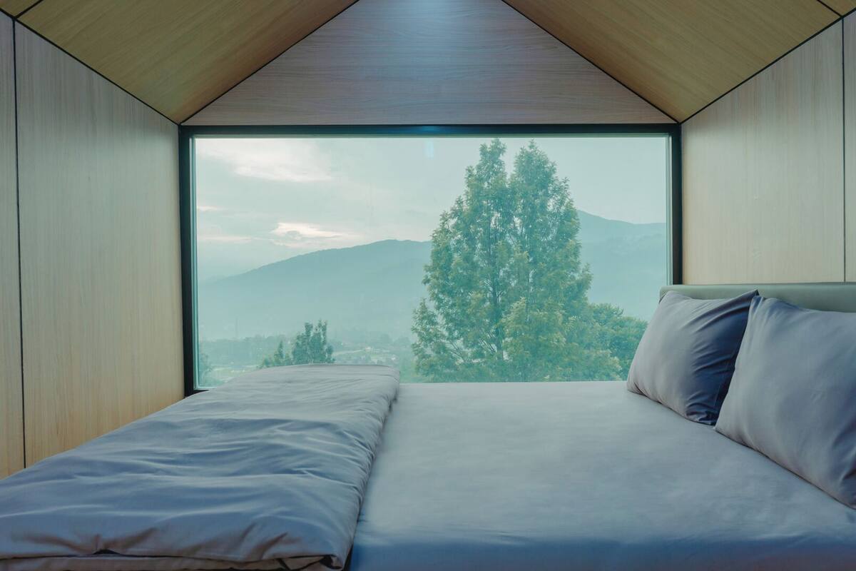 Quarto com uma janela grande, uma cama e vista para uma árvore e montanhas.
