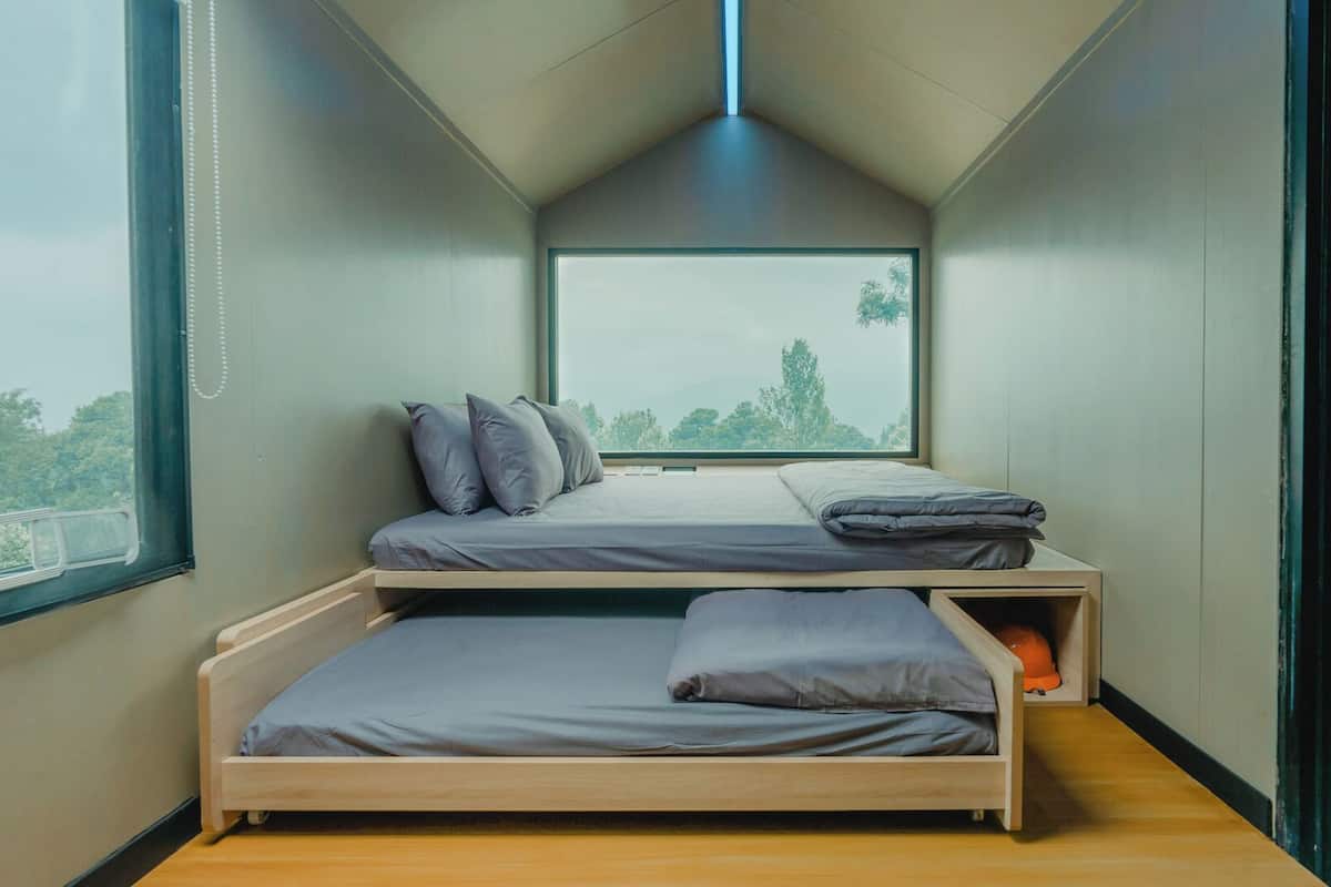 Quarto compacto com beliche e cama auxiliar, janelas amplas e vista para árvores.
