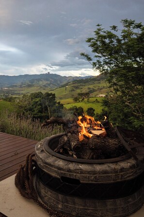 Uma lareira de pedra com fogo aceso, cercada por um deck de madeira e com vista para uma paisagem pitoresca com colinas e árvores.