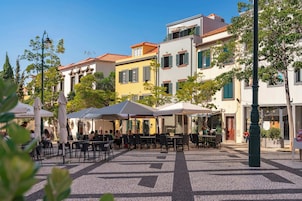 Uma área de café ao ar livre, com mesas e cadeiras, rodeada por edifícios com varandas e janelas.