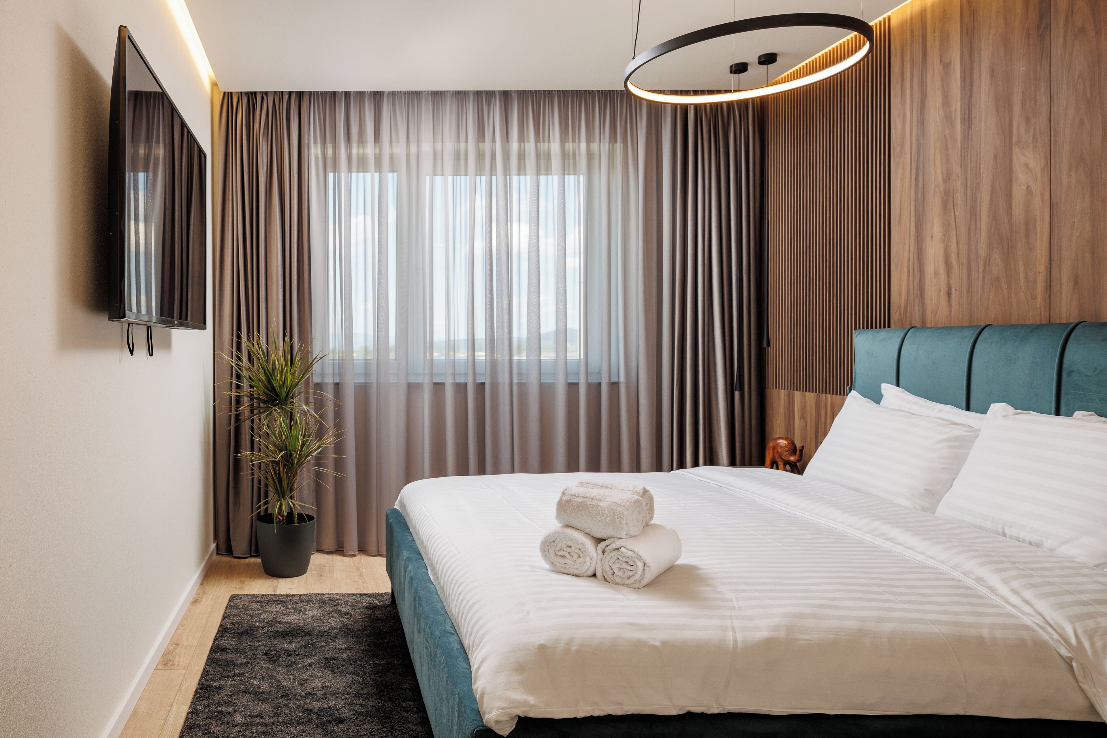 Quarto de hotel moderno com uma cama grande, televisão de tela plana, planta em vaso e vista da cidade pela janela.