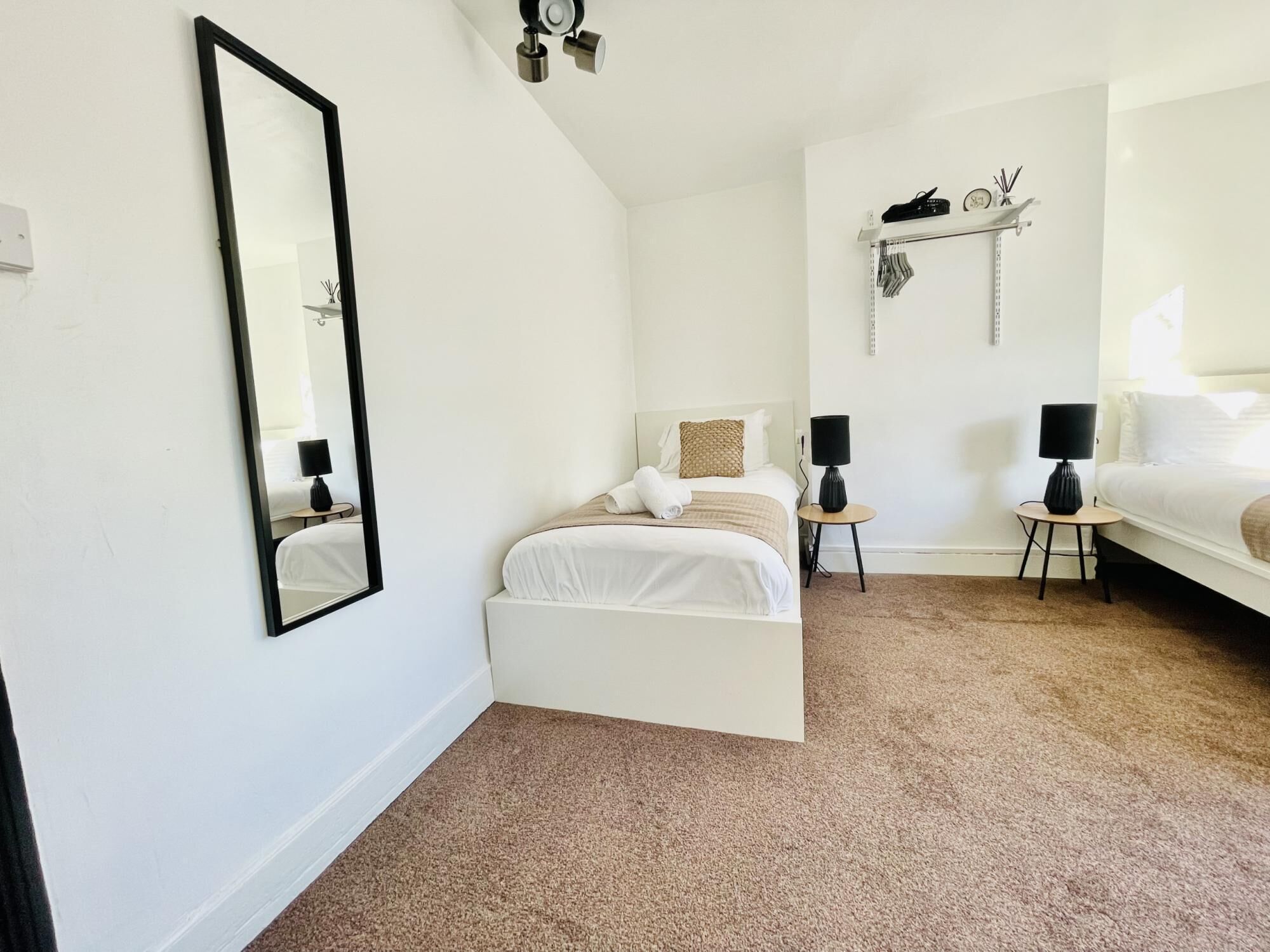 Quarto de hotel moderno com uma cama grande, mesa de cabeceira com abajur, mesinha lateral e prateleira na parede com objetos decorativos.