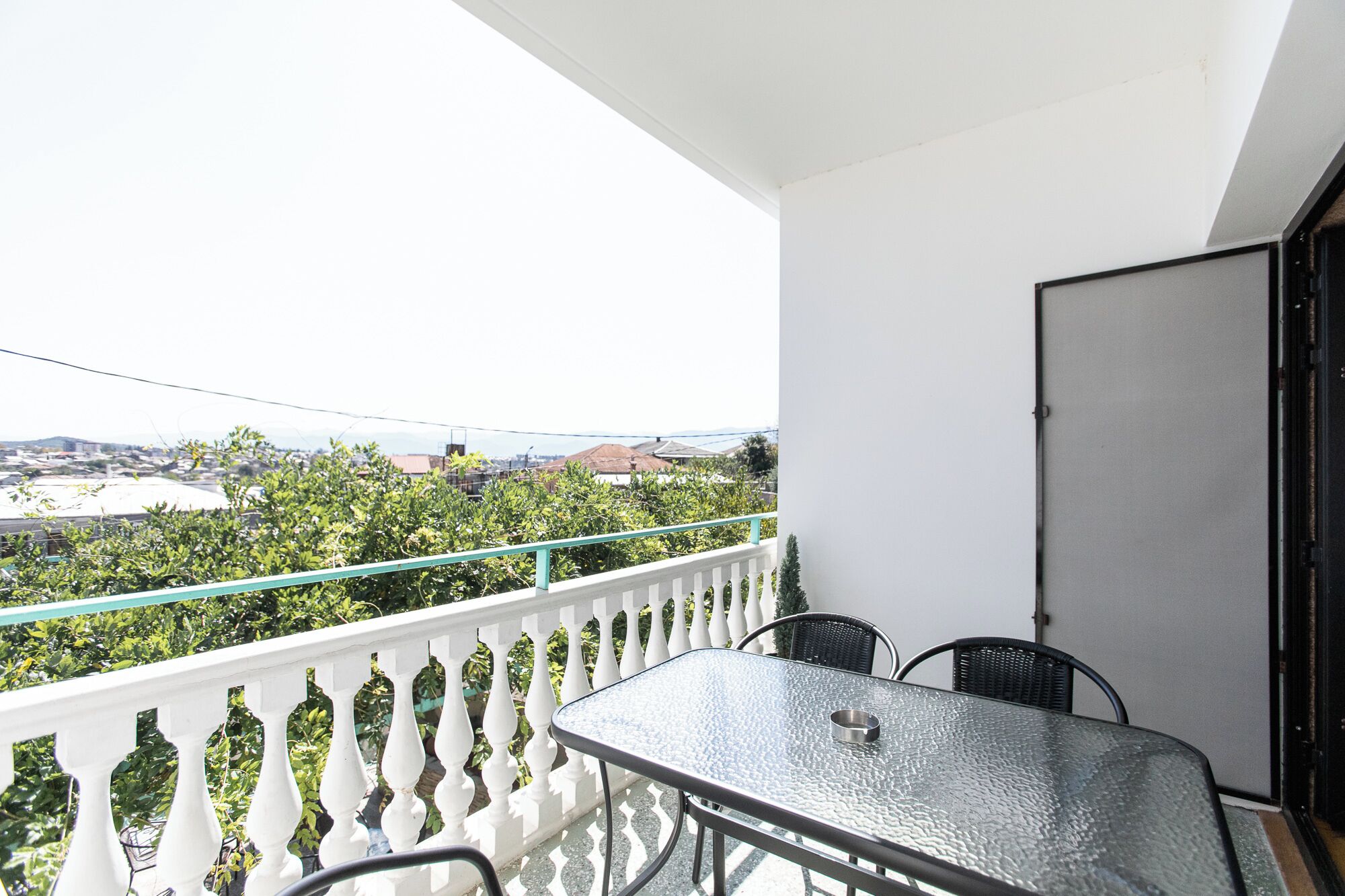 Varanda com mesa e cadeiras de vidro, balaustrada branca e vista para a cidade.