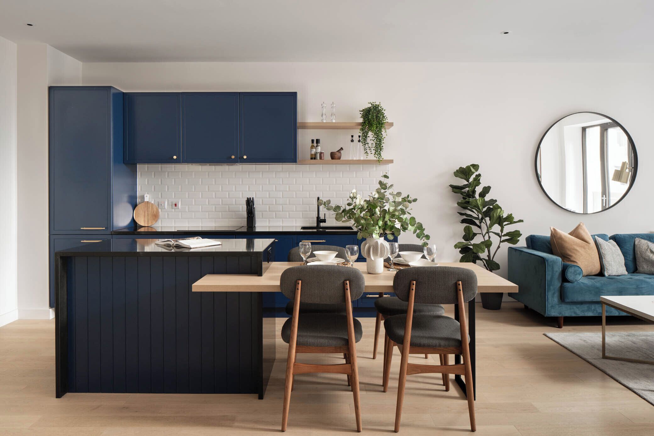 Uma cozinha moderna com armários azul escuro, painel de tijolos brancos e uma ilha central com cadeiras. Há um sofá azul e um espelho redondo na parede.
