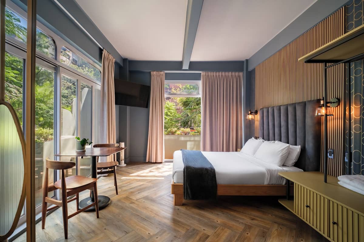 Quarto de hotel moderno com uma cama grande, piso de madeira, área de jantar e vista para o verde lá fora.