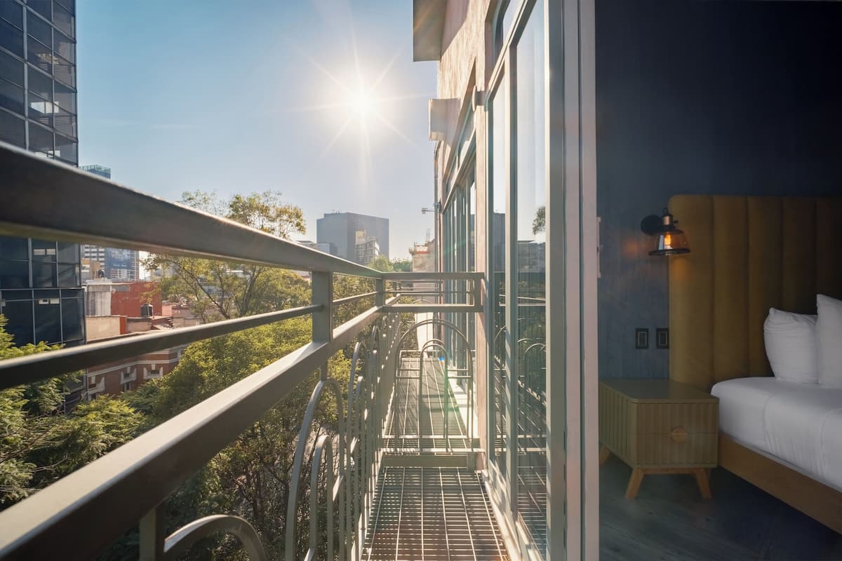 Varanda com grades de metal dando vista para a cidade, uma cama com mesa de cabeceira de madeira e um abajur fixado na parede.