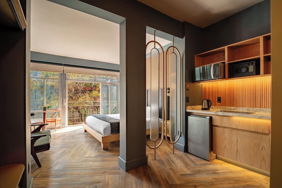 Quarto de hotel moderno com piso de madeira, cama, escrivaninha, cadeira, área de cozinha com micro-ondas e cafeteira, e uma grande janela com vista para árvores.