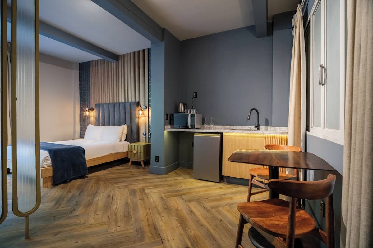Quarto de hotel compacto com cama, mesa de cabeceira e uma pequena cozinha.