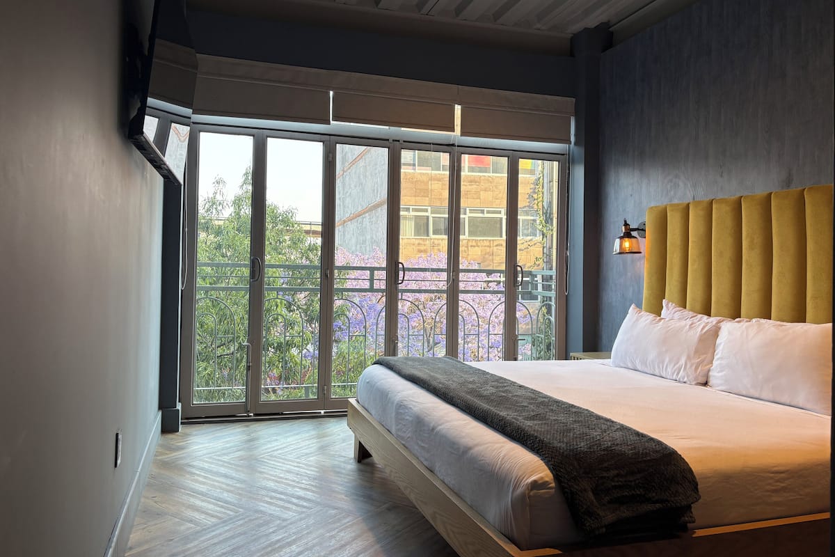Quarto com uma janela grande, cama com roupa de cama branca e cabeceira amarela.