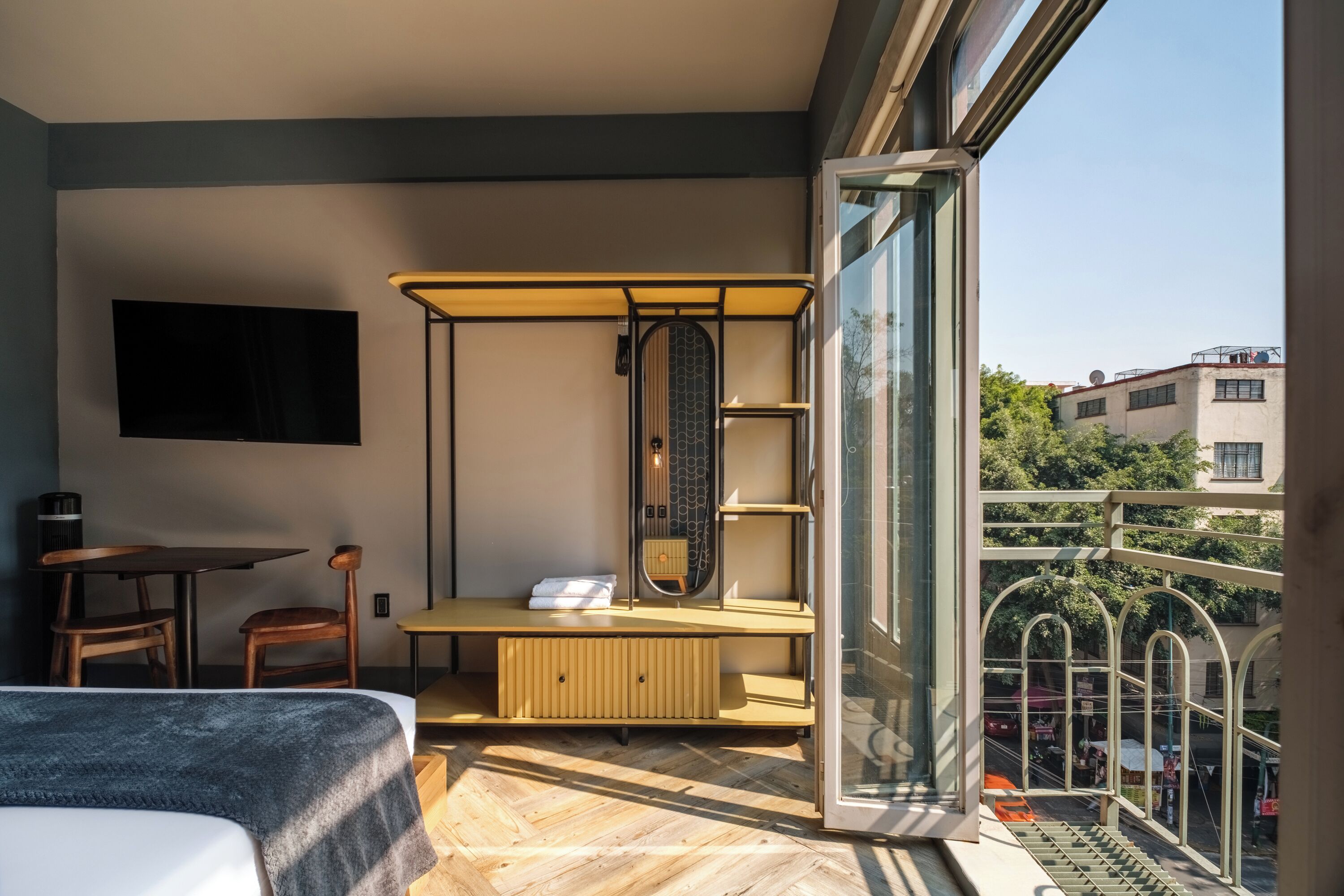 Quarto moderno com cama, escrivaninha, cadeira, televisão e uma grande janela com vista para edifícios e árvores.