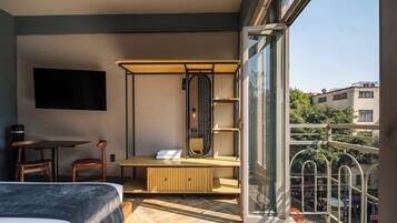 Quarto moderno com cama, escrivaninha, cadeira, televisão e uma grande janela com vista para edifícios e árvores.