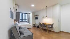 Quarto de hotel moderno com sofá, mesa de jantar com cadeiras, televisão na parede e uma janela grande com cortinas.