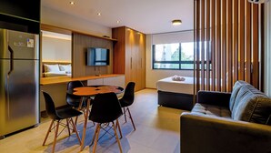 Quarto de hotel moderno com cozinha compacta, área de refeições com mesa e cadeiras redondas, um sofá e uma TV na parede.