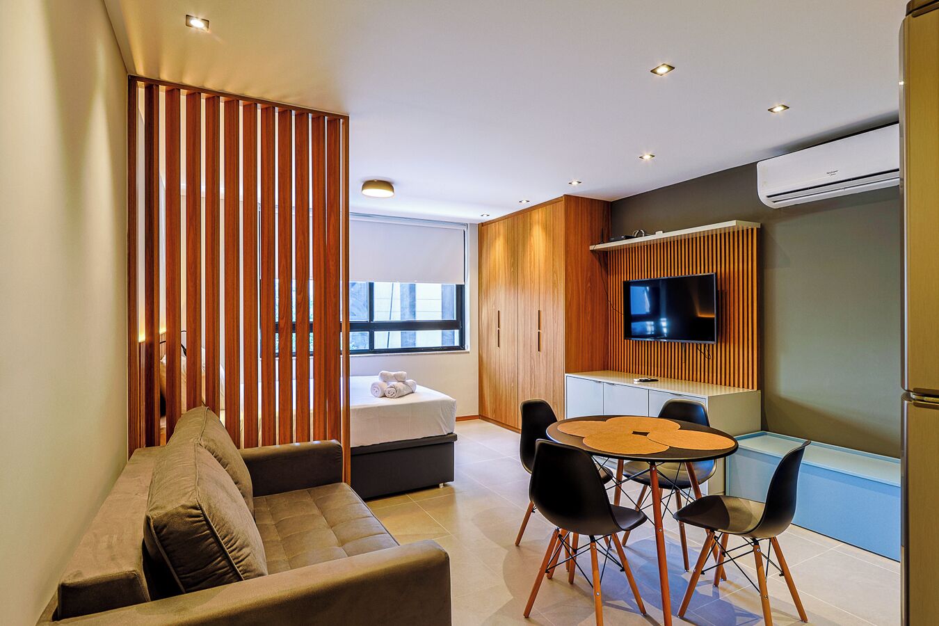 Quarto de hotel moderno com sofá, mesa de jantar com cadeiras, TV fixada na parede e aparelho de ar condicionado.