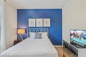 Quarto de hotel com cama, televisão, mesa de cabeceira, abajur e três quadros na parede.