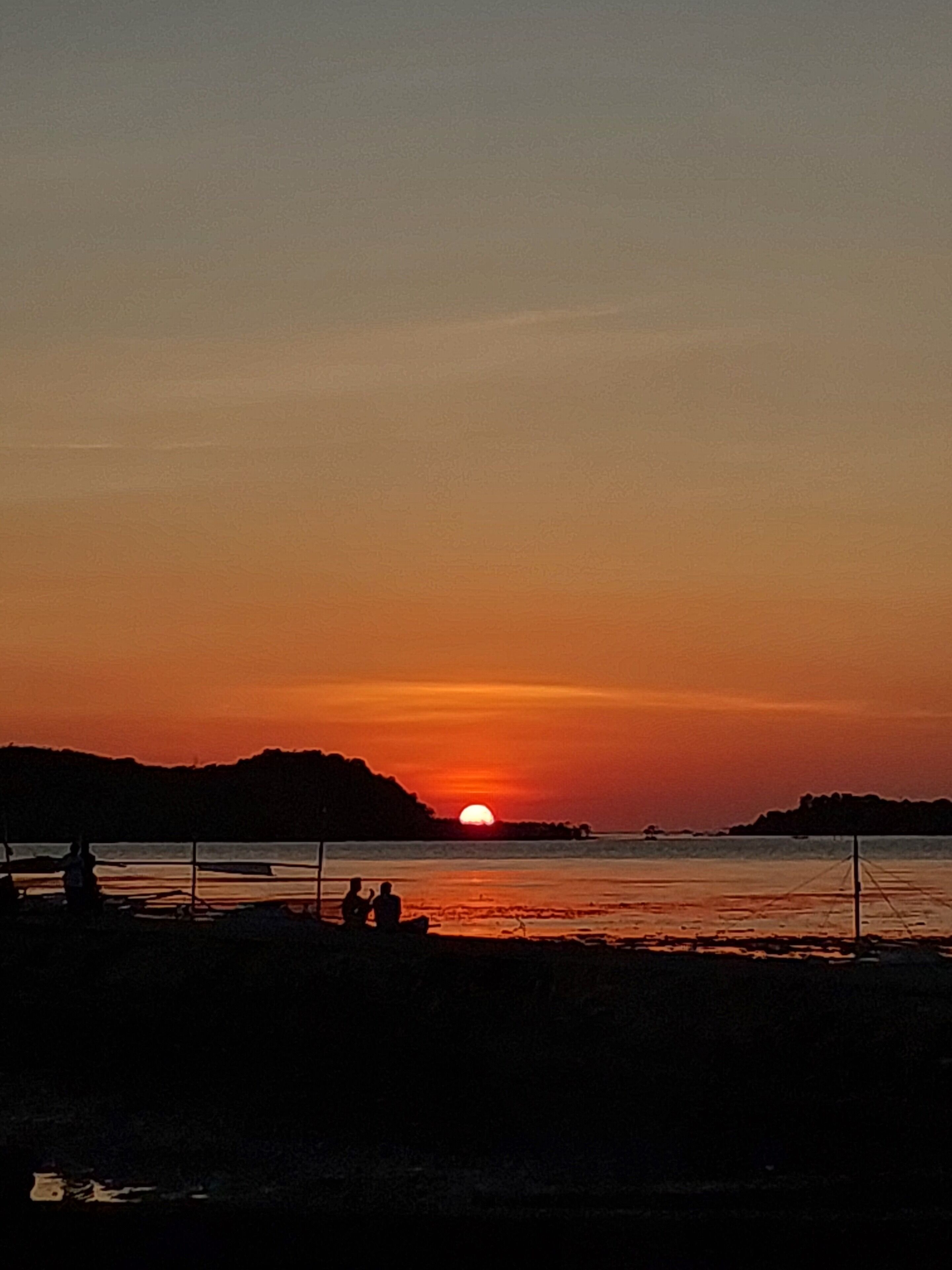 Pôr do sol sobre a água, com silhuetas de pessoas e uma terra ao longe.