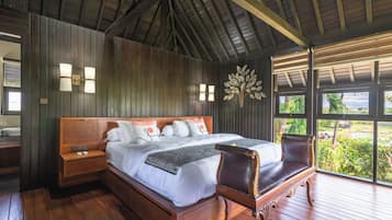 Quarto espaçoso com uma cama grande, cabeceira de madeira, mesas de cabeceira e vista para vegetação exuberante através de janelas amplas.