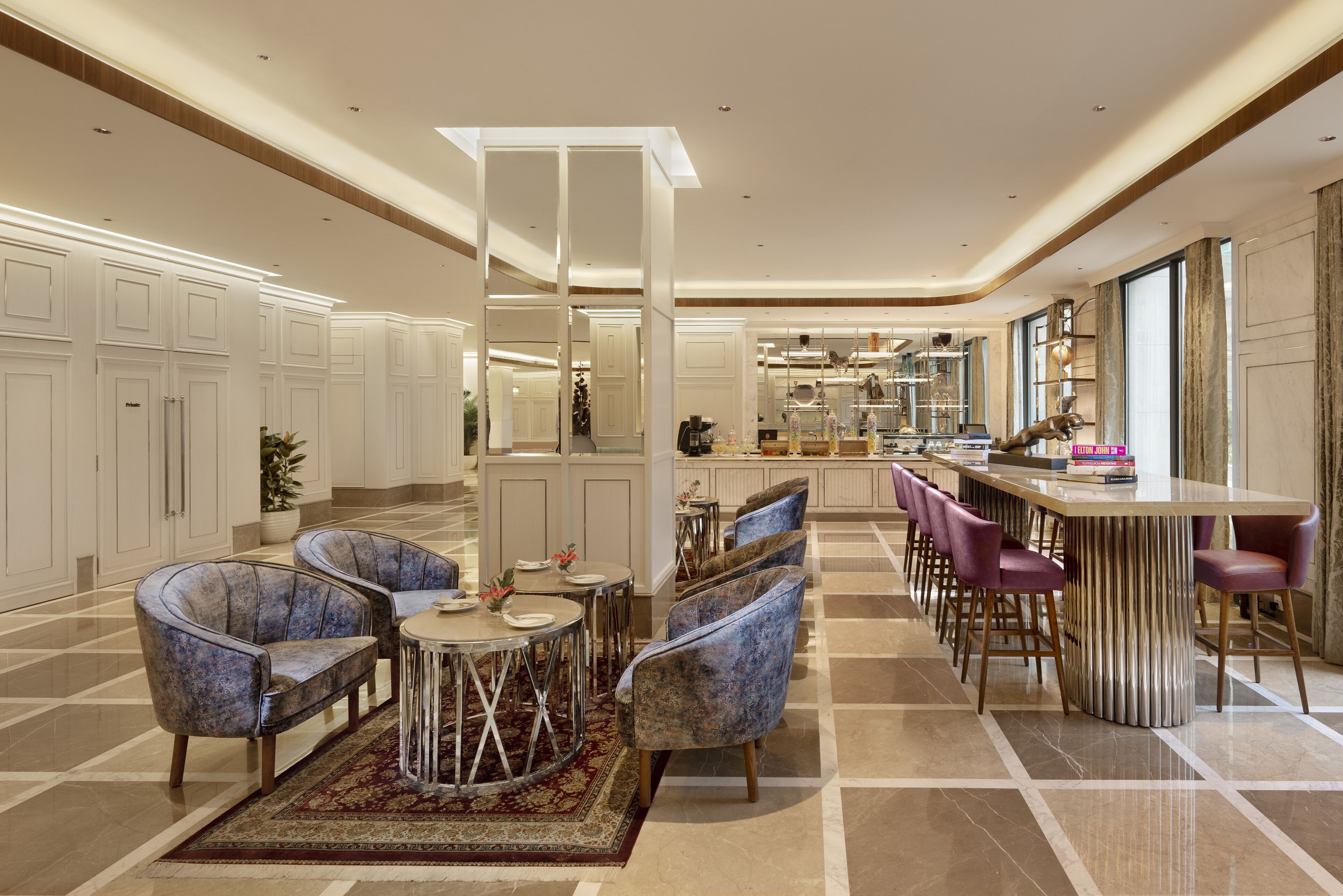 Um lobby de hotel espaçoso e elegantemente decorado, com área de estar central, bar e vista para a cozinha ao fundo.