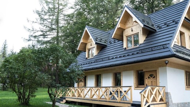 Uma casa de madeira de dois andares, com telhado inclinado, varanda de madeira e um caminho de cascalho que leva à entrada.