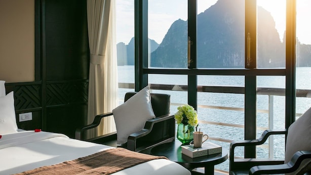Quarto de hotel com uma grande janela que oferece vista para o mar e montanhas. Há uma cama, uma cadeira e uma pequena mesa com um vaso de flores e um livro intitulado 