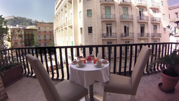 Uma varanda com duas cadeiras, uma pequena mesa, um vaso de planta e vista para um edifício de vários andares com varandas.