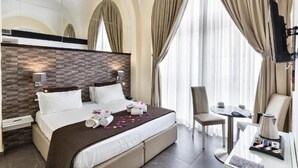 Quarto de hotel com uma cama grande, uma televisão de tela plana, uma mesinha com cafeteira e um espaço de estar com uma poltrona.
